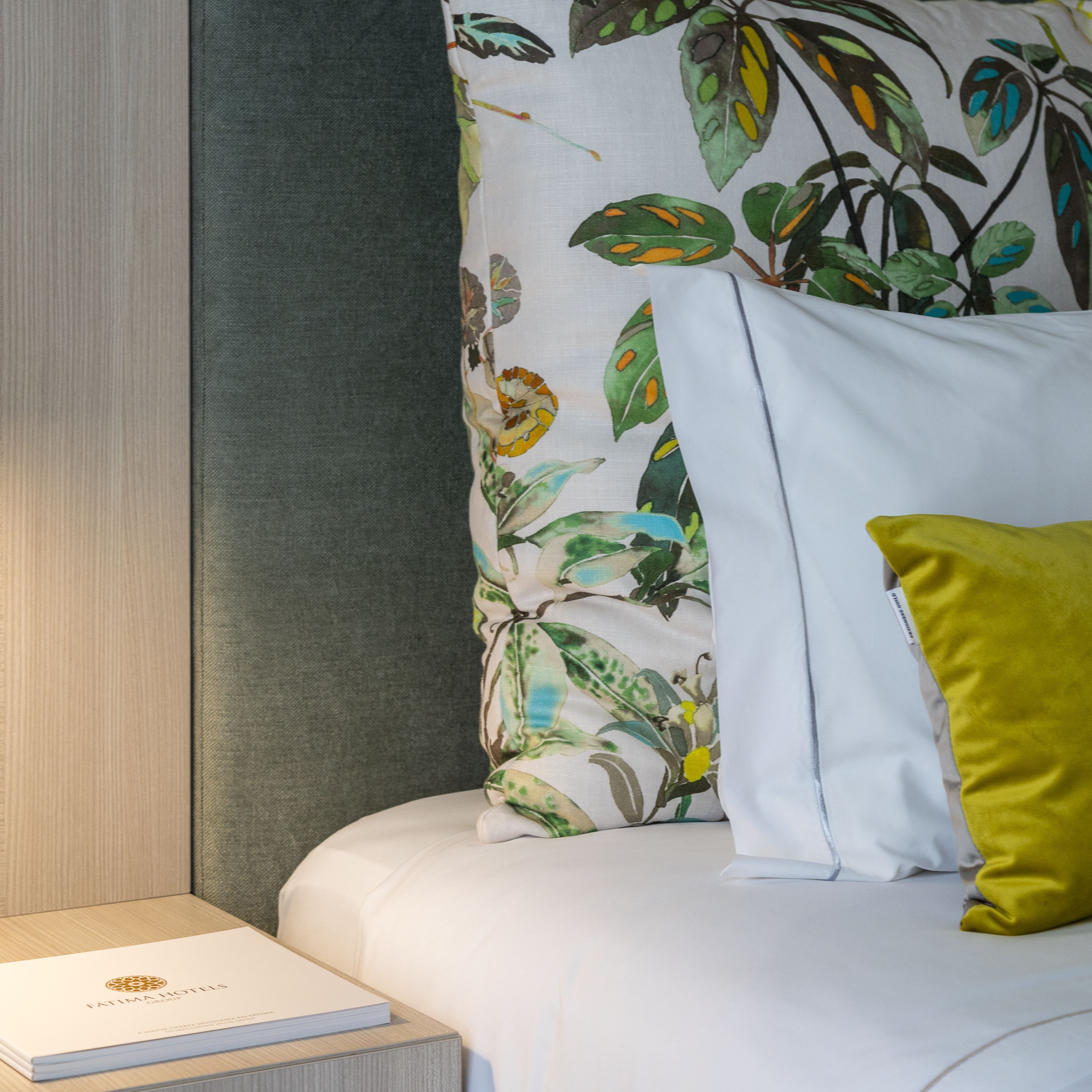 Cama bem arrumada com travesseiro branco e um travesseiro de detalhe amarelo, cortina com padrão floral e uma pilha de livros em uma mesa de cabeceira.