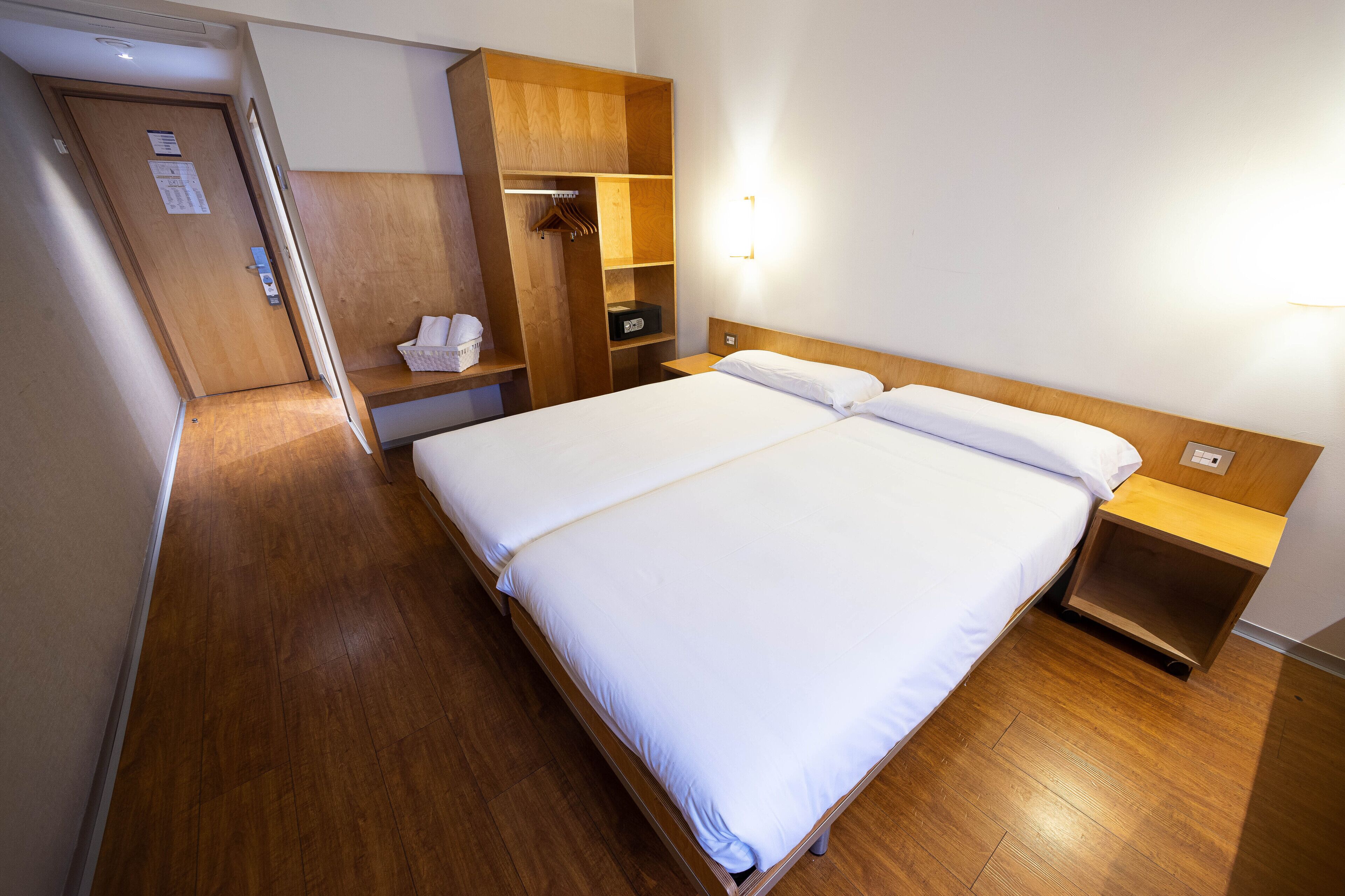 Quarto de hotel com uma cama grande, guarda-roupa de madeira, mesas de cabeceira e luminária fixada na parede.
