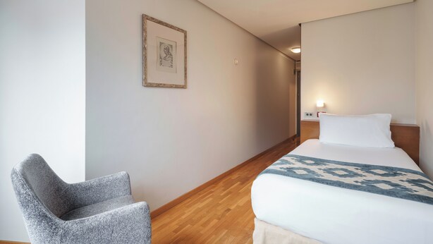 Quarto de hotel com uma cama, uma cadeira, um quadro na parede e um corredor que dá para outro quarto.