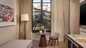 Quarto de hotel com cama, mesa redonda com um vaso de flores, cadeira, abajur, janela com cortinas e um quadro na parede.
