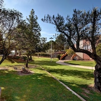 Um parque com um calçadão pavimentado, árvores e um edifício com fachada vermelha ao fundo.