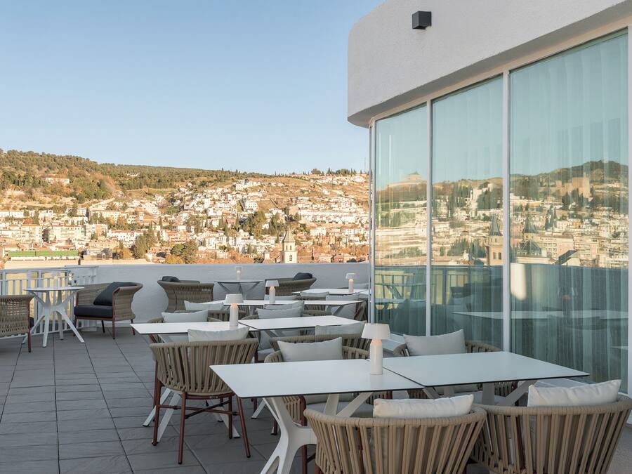 Um terraço no topo com mesas e cadeiras de vime, com vista para uma cidade numa colina.