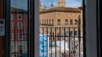 Vista de varanda com guarda-corpo em ferro forjado, com vista para um edifício histórico com uma torre imponente e uma praça com uma estátua.