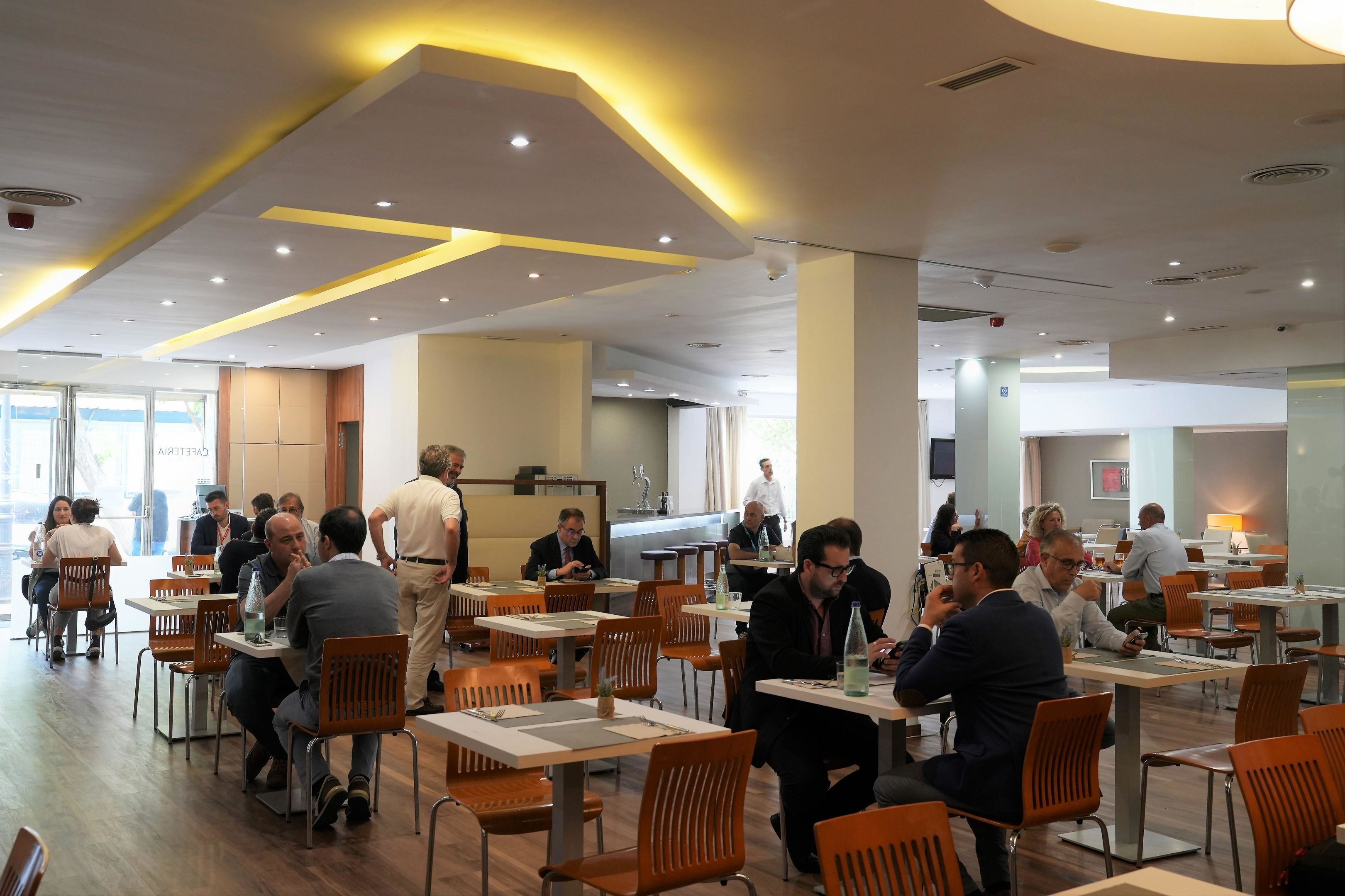 Uma sala de jantar ampla com várias mesas e cadeiras, cozinha ao fundo e um design de teto moderno.