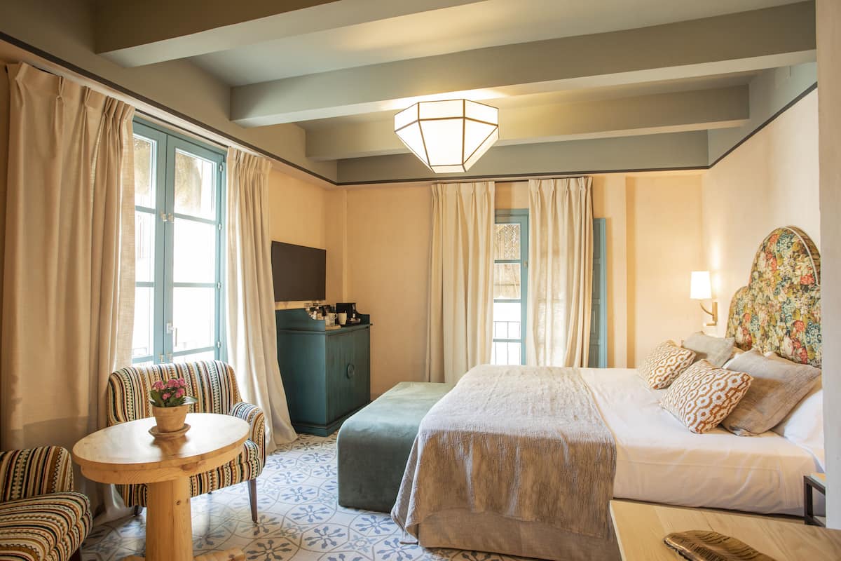 Quarto de hotel com cama, escrivaninha, cadeira, cômoda e janela com cortinas.
