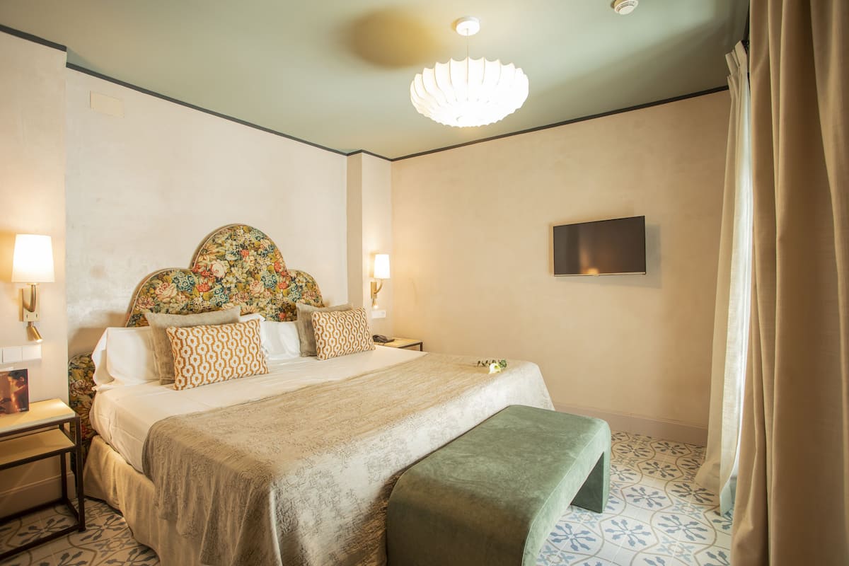 Quarto de hotel com uma cama grande, mesa de cabeceira, abajur, painel decorativo de cabeceira, TV de tela plana e cortina.