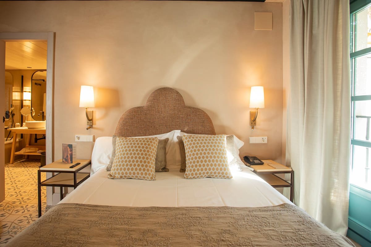 Quarto de hotel com uma cama grande, duas mesas de cabeceira, um criado-mudo, um banheiro visível por uma porta aberta e uma janela com cortinas.