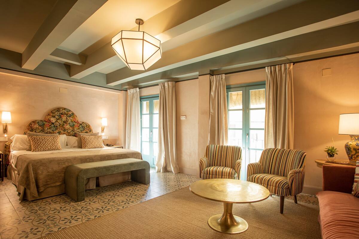 Quarto de hotel com uma cama, um banco, duas poltronas, uma mesa redonda e um lustre.