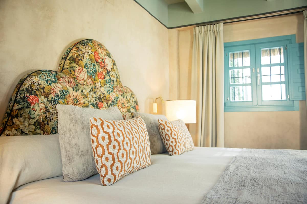 Cama com almofadas de cabeceira estampadas, luminária de apoio e janela com persianas azuis.
