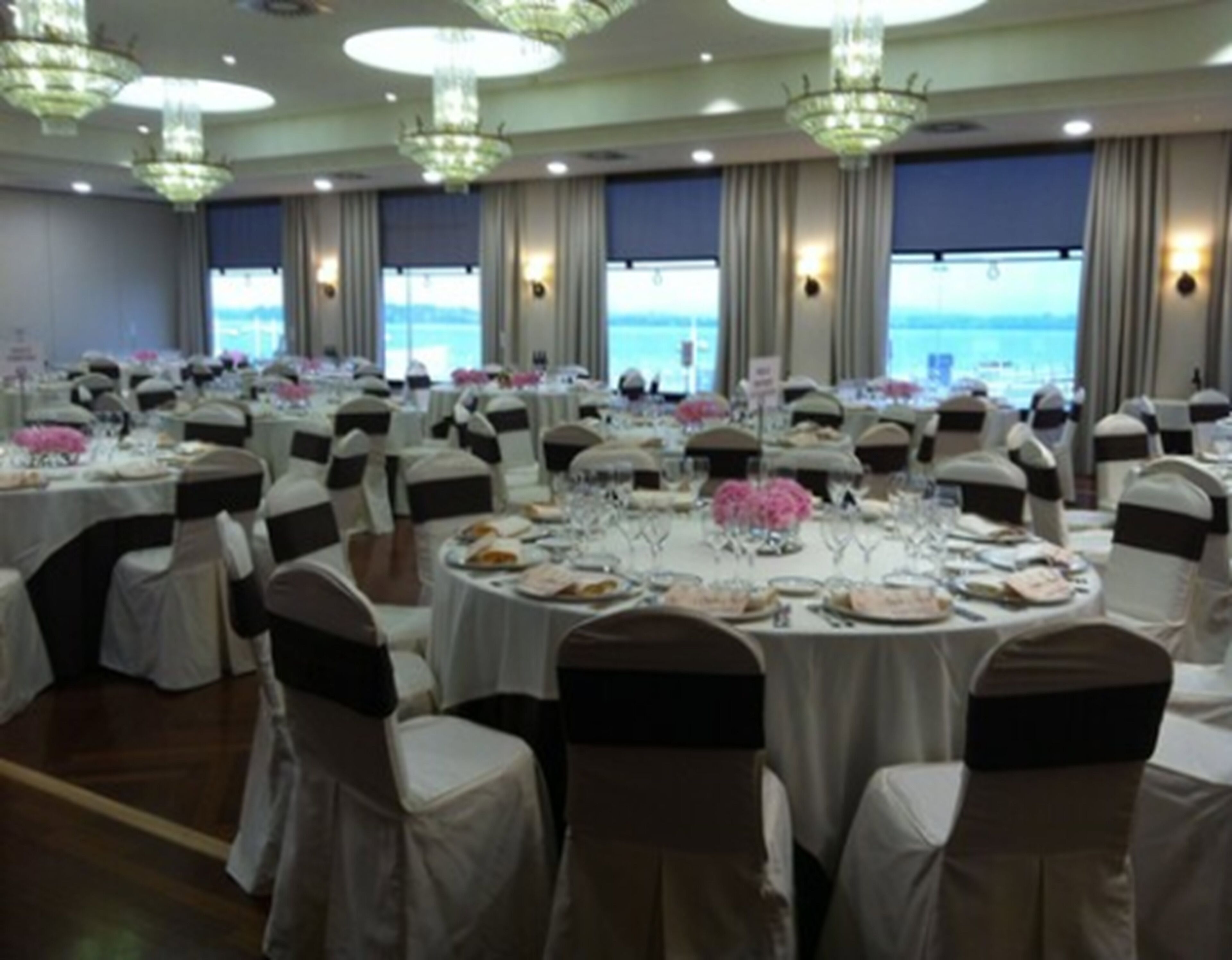 Uma sala de banquetes com mesas redondas preparadas para um evento formal, grandes janelas com vista e lustres.