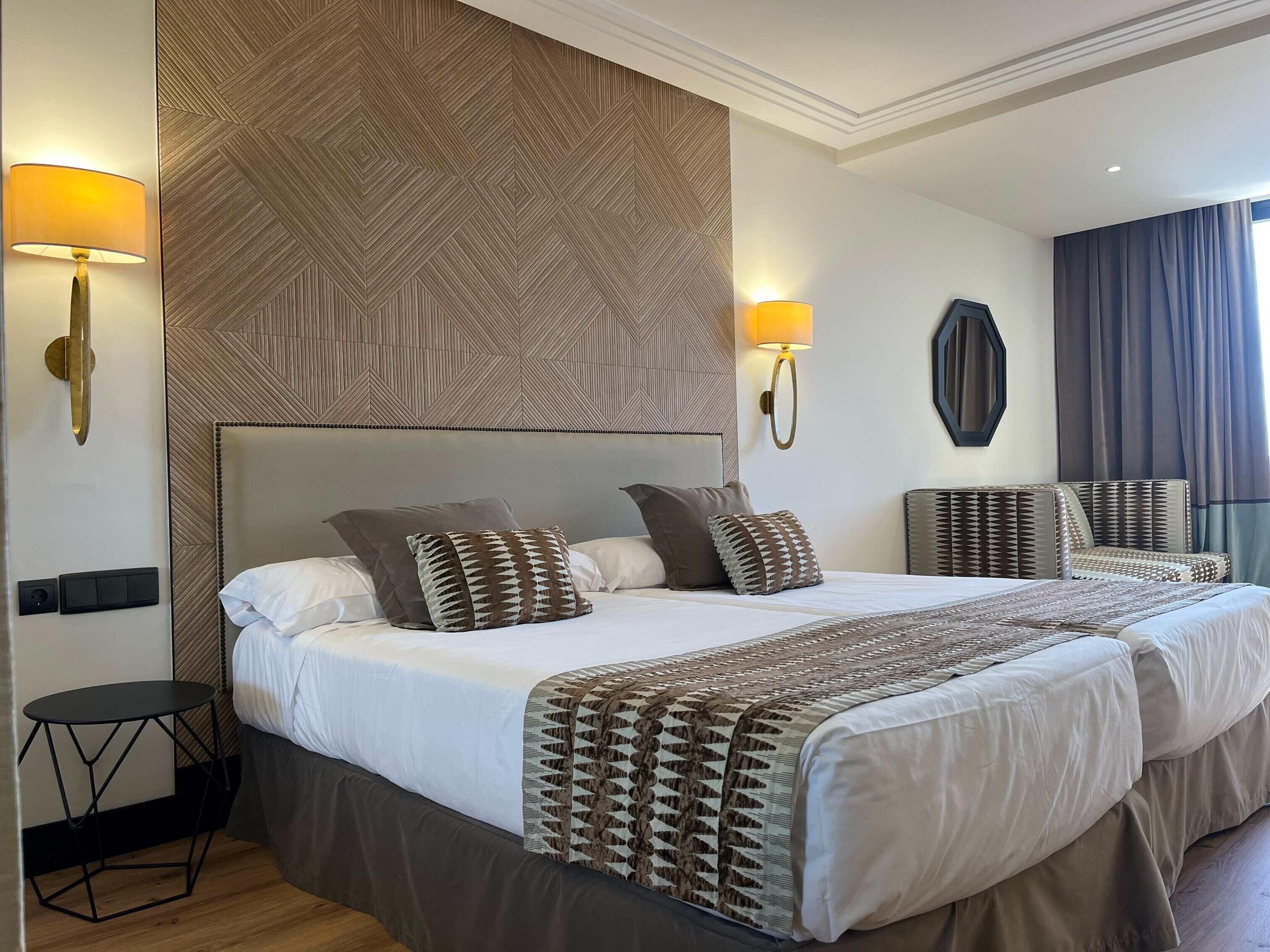 Quarto de hotel com uma cama grande, duas mesas de cabeceira, uma luminária na parede e uma janela com cortinas.