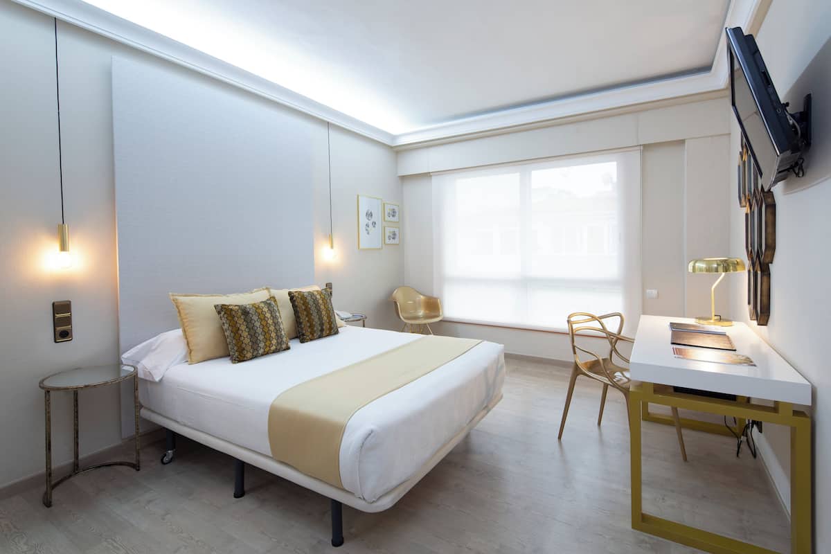 Quarto de hotel moderno com cama, mesa de cabeceira, cadeira, escrivaninha e TV na parede.
