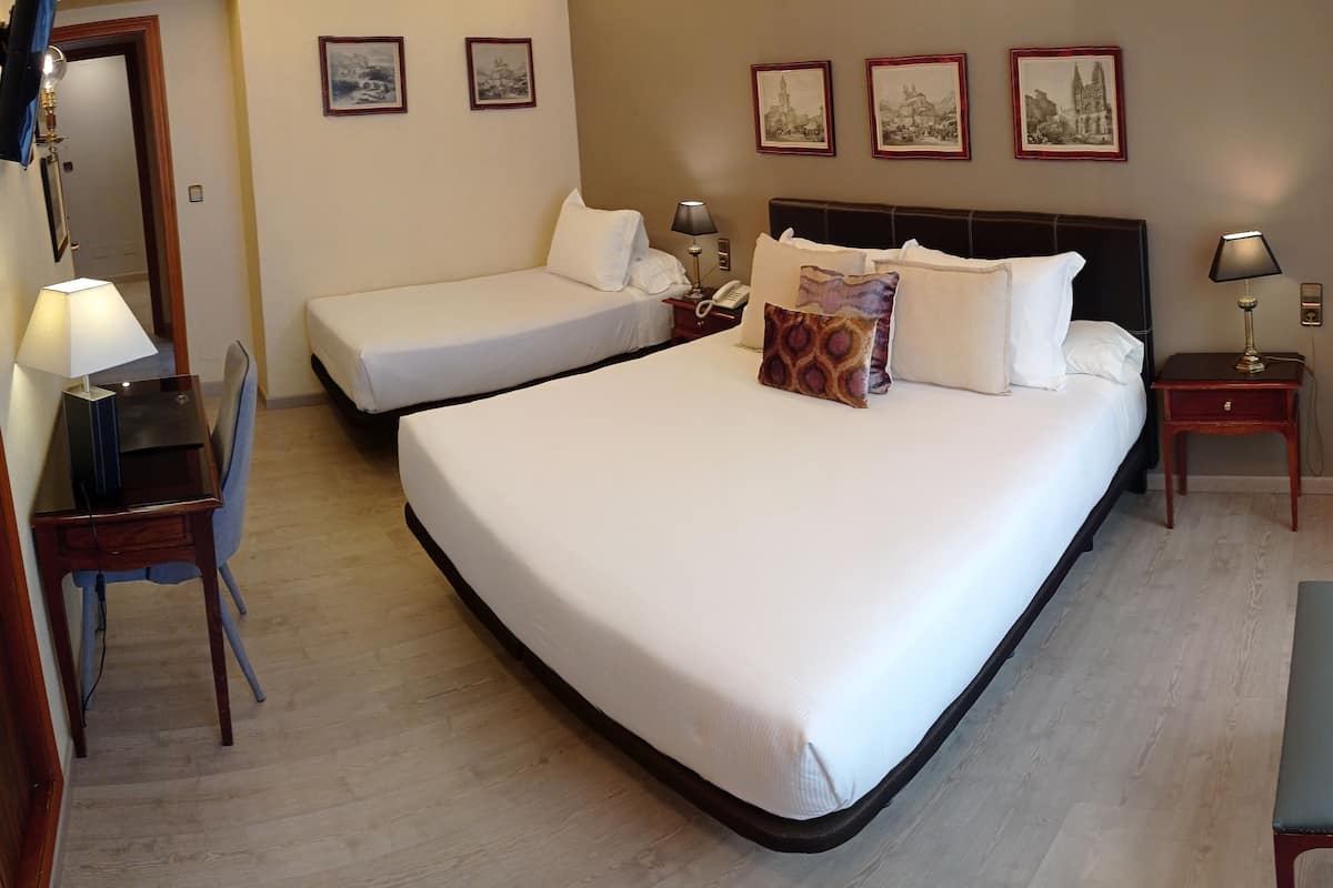 Quarto de hotel com duas camas de solteiro, uma escrivaninha com cadeira, mesa de cabeceira com abajur e um banco.
