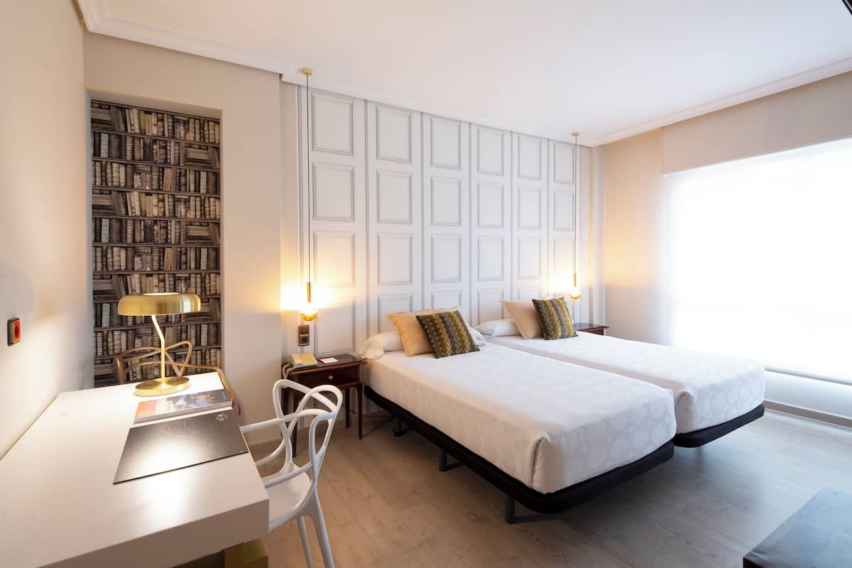 Quarto de hotel com duas camas, uma escrivaninha, uma cadeira, estante de livros e uma janela grande.