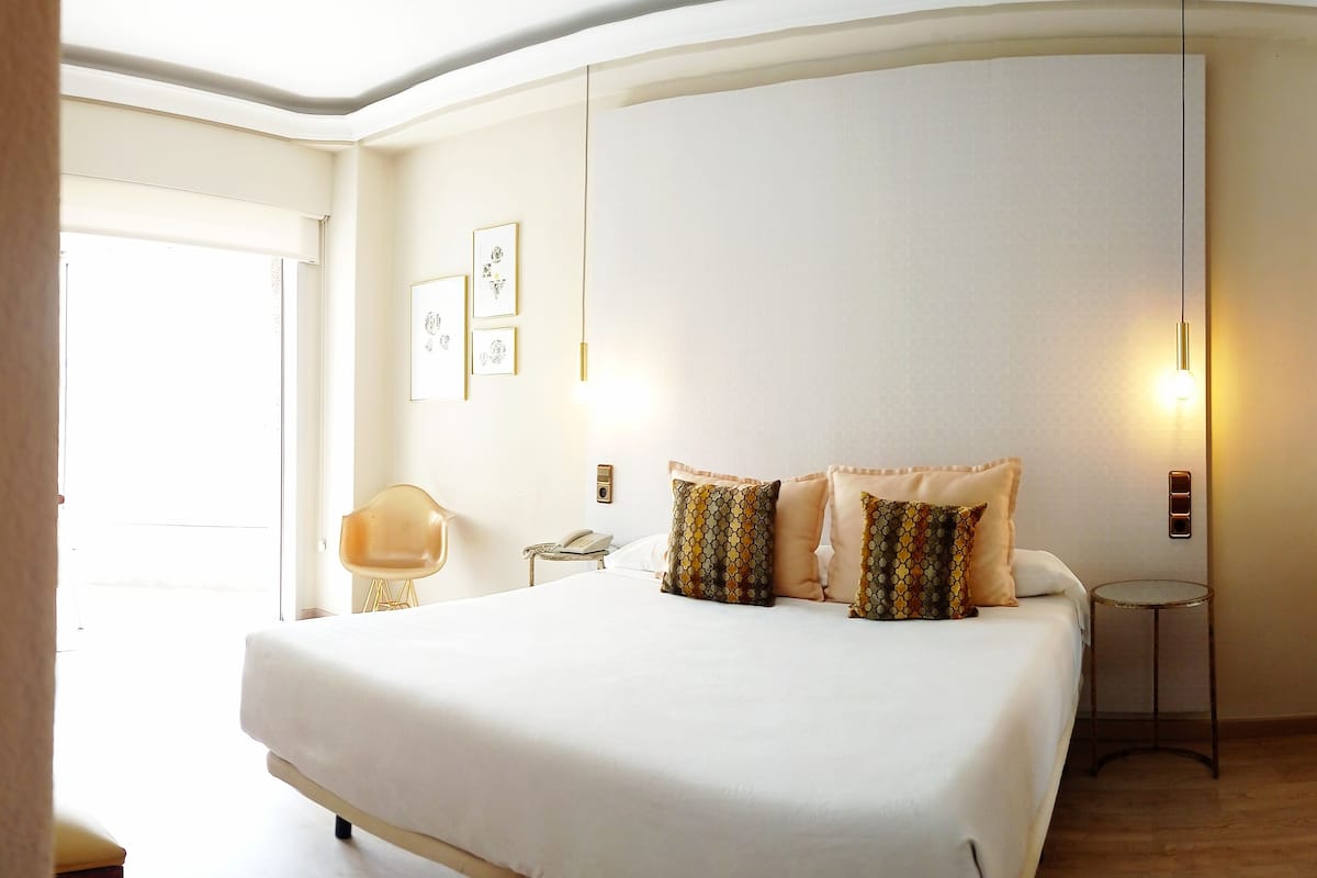 Quarto de hotel com uma cama grande, mesas de cabeceira, uma cadeira e uma pequena mesa.