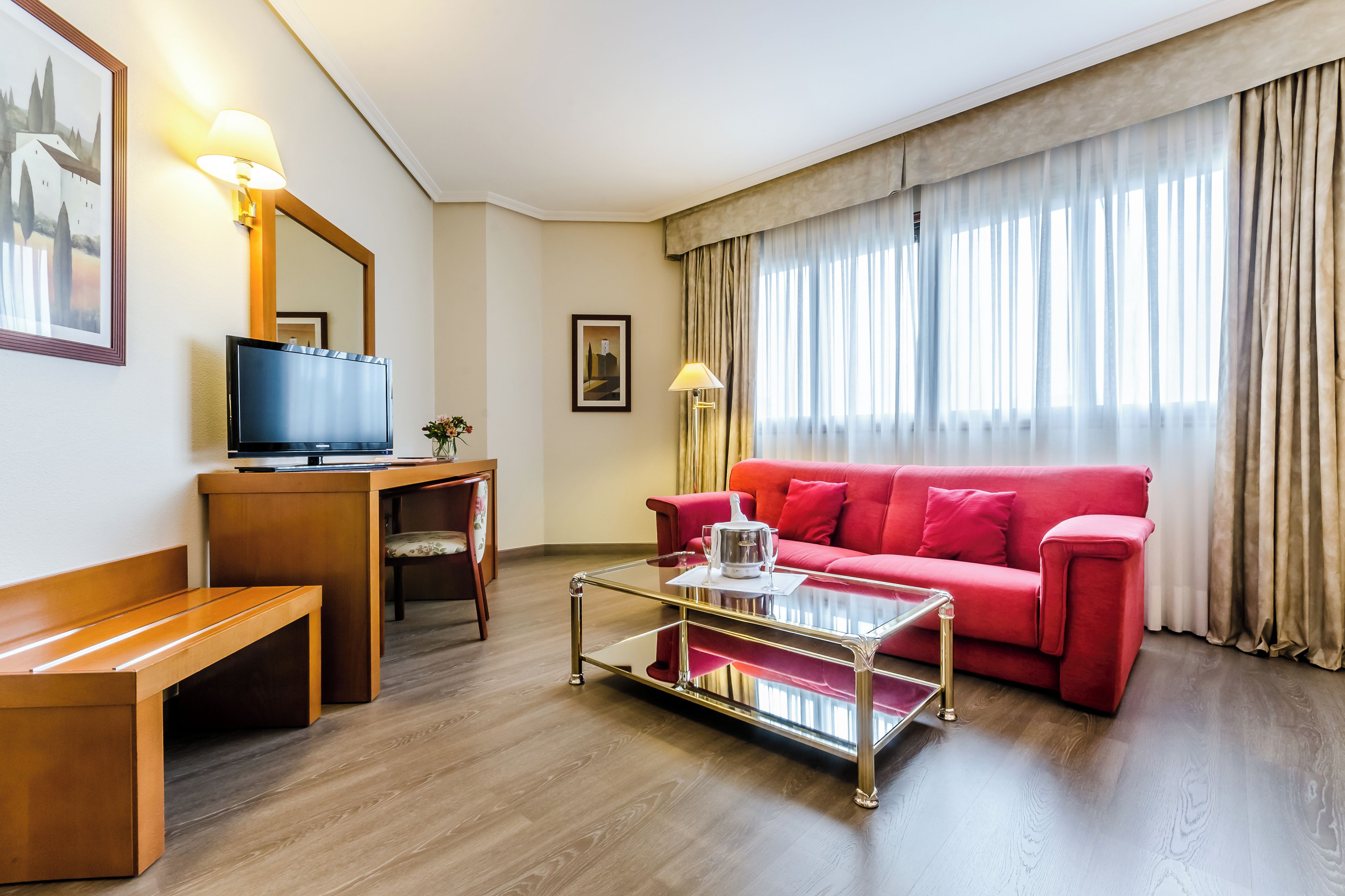 Quarto de hotel com televisão de tela plana, sofá vermelho, mesa de centro de vidro e escrivaninha de madeira.