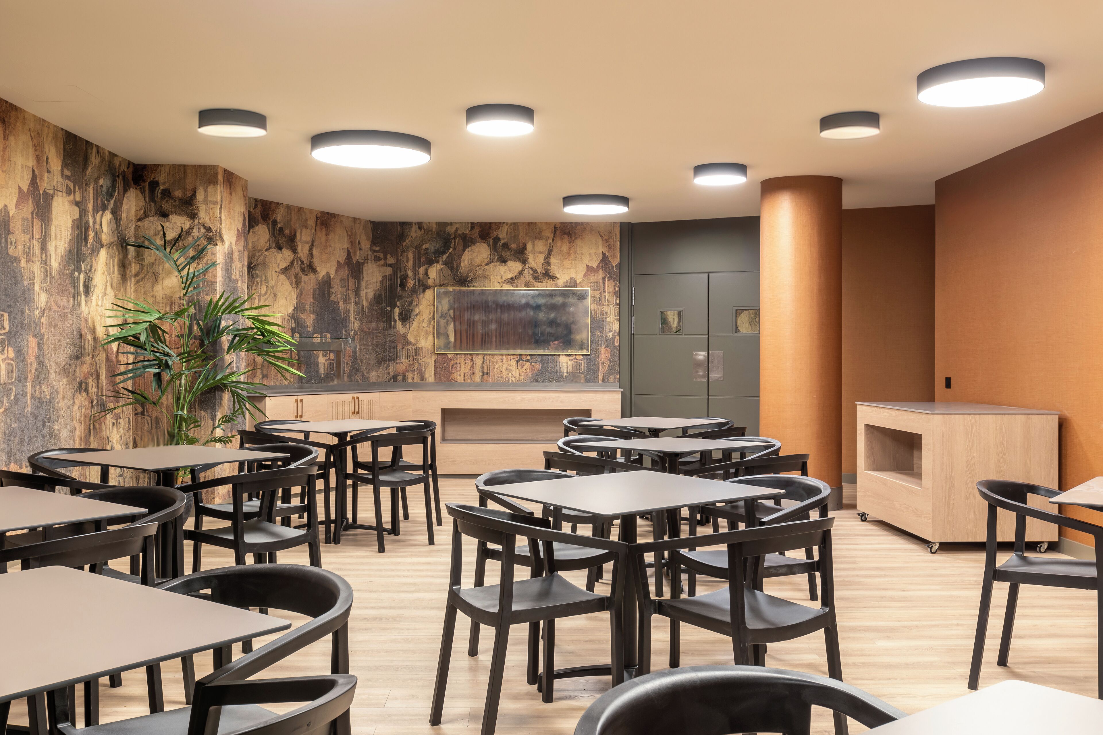 Uma área de refeições moderna, com várias mesas e cadeiras, um painel de madeira decorativo e um balcão de recepção.