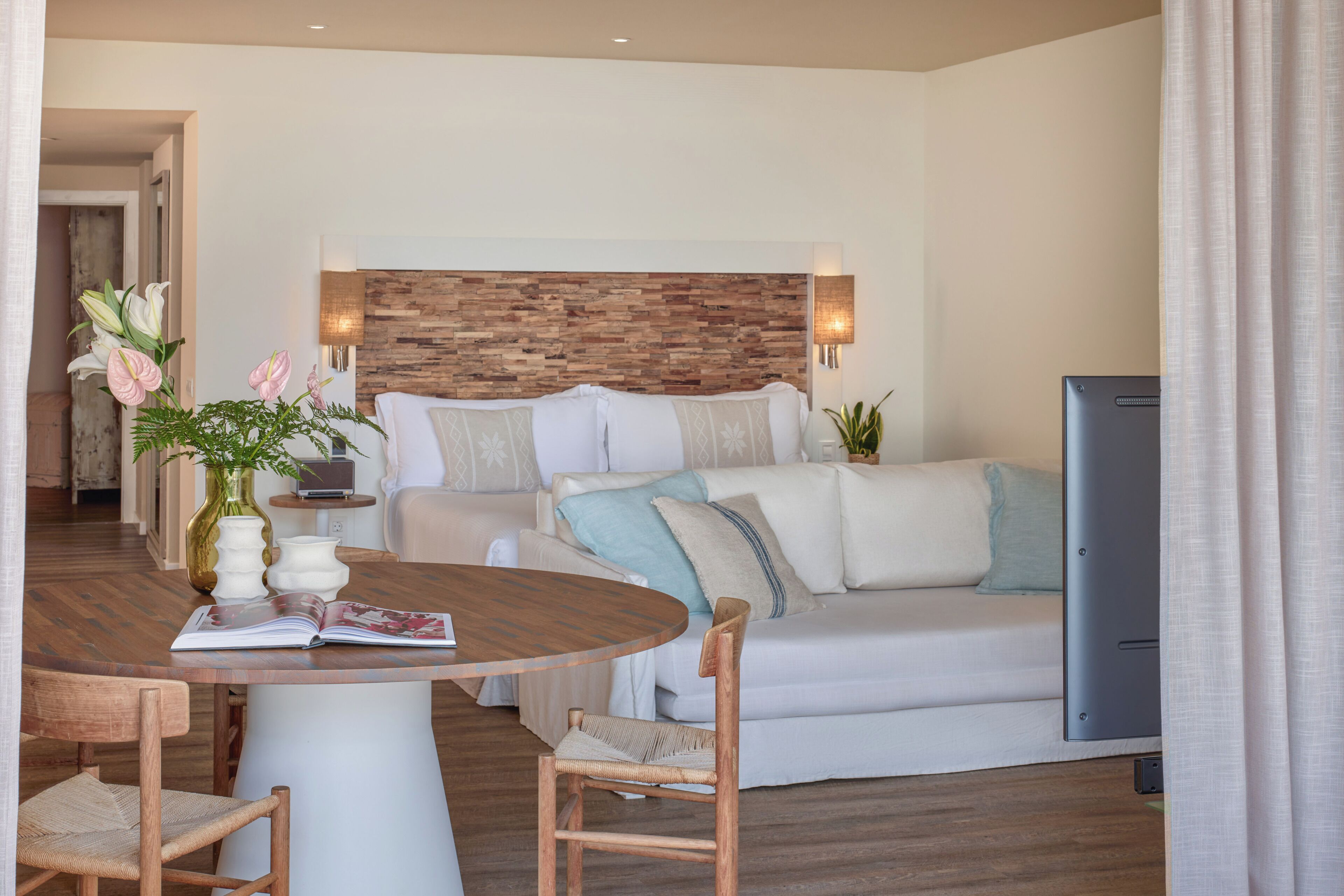 Quarto de hotel moderno com cabeceira de madeira, sofá branco, mesa de jantar com cadeiras e televisão de tela plana.