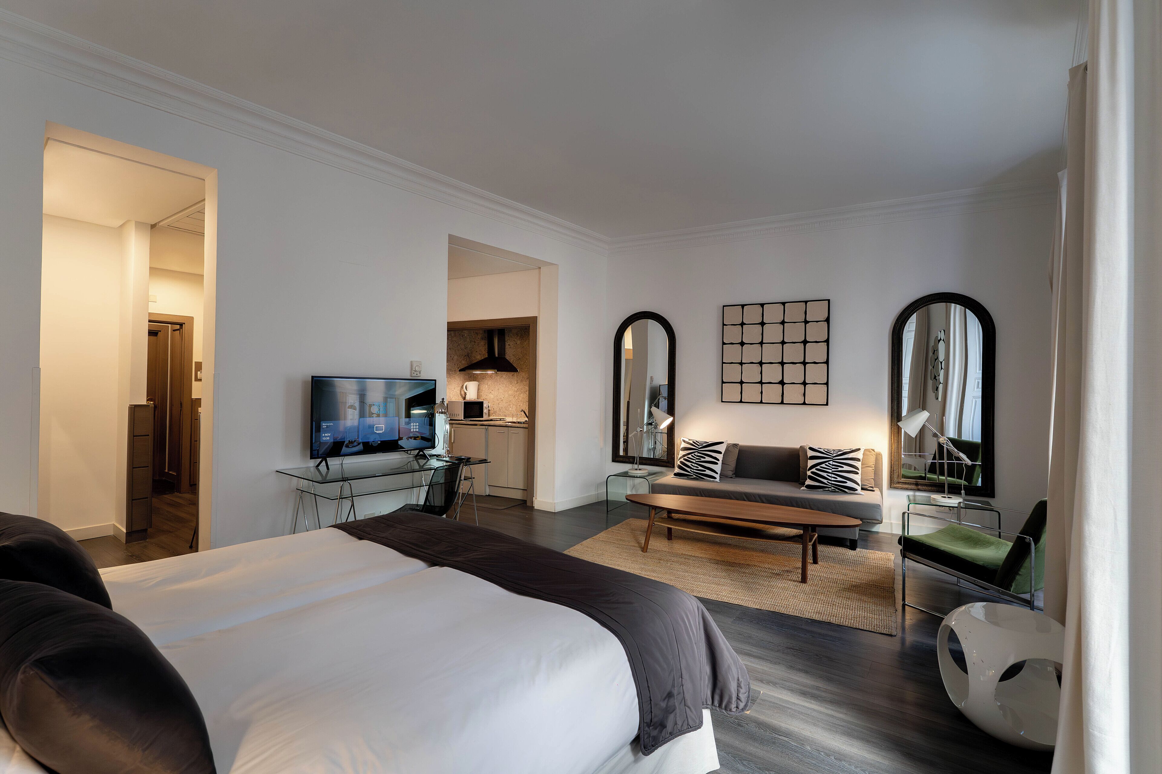 Quarto de hotel moderno com uma cama grande, uma escrivaninha com computador, um sofá e dois espelhos.