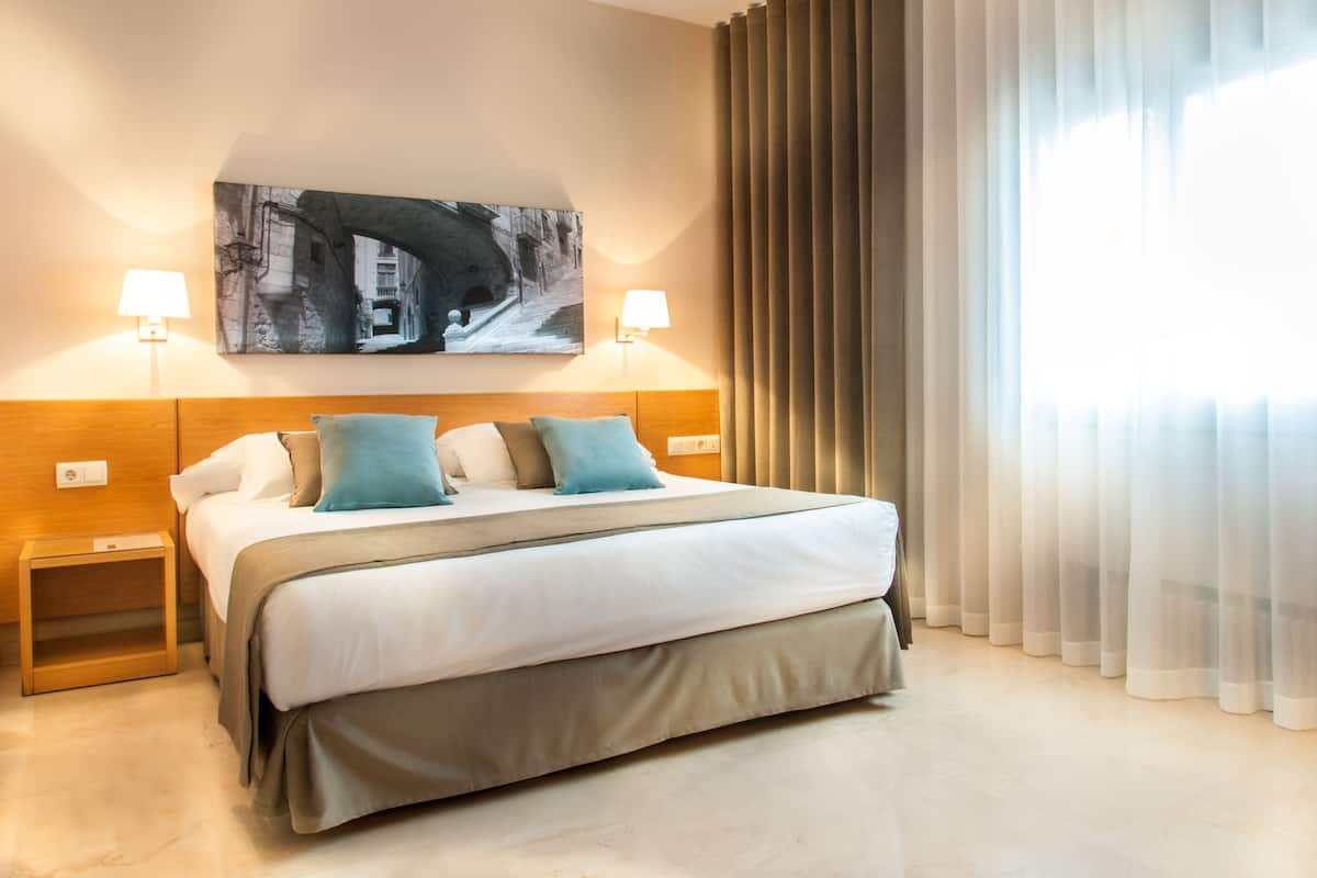 Quarto de hotel com cama, duas luminárias de cabeceira, um quadro na parede e uma janela com cortinas.