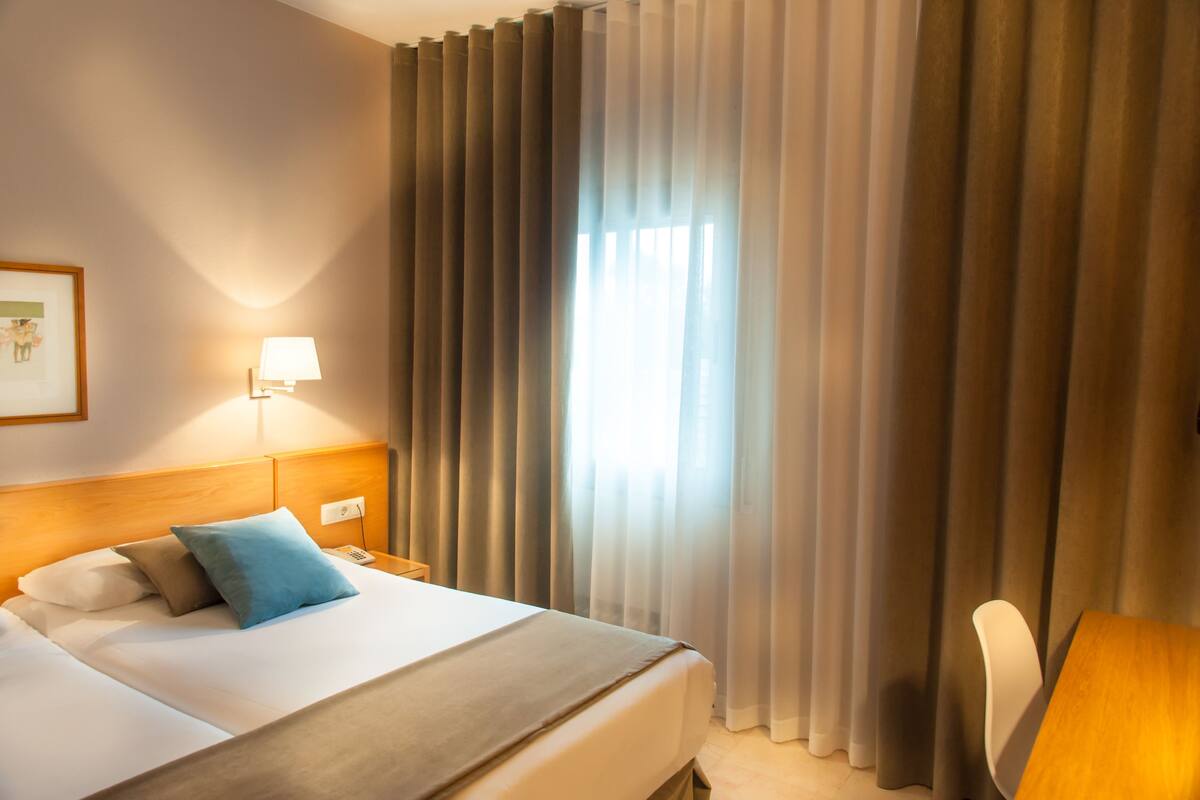 Quarto de hotel com duas camas, uma mesa de cabeceira com abajur, uma cadeira e uma janela com cortinas.