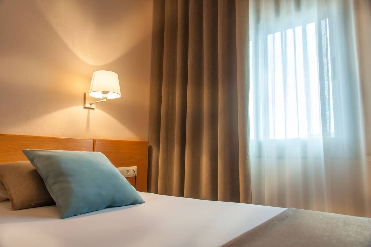 Quarto de hotel com cama, luminária na parede e janela com cortinas.