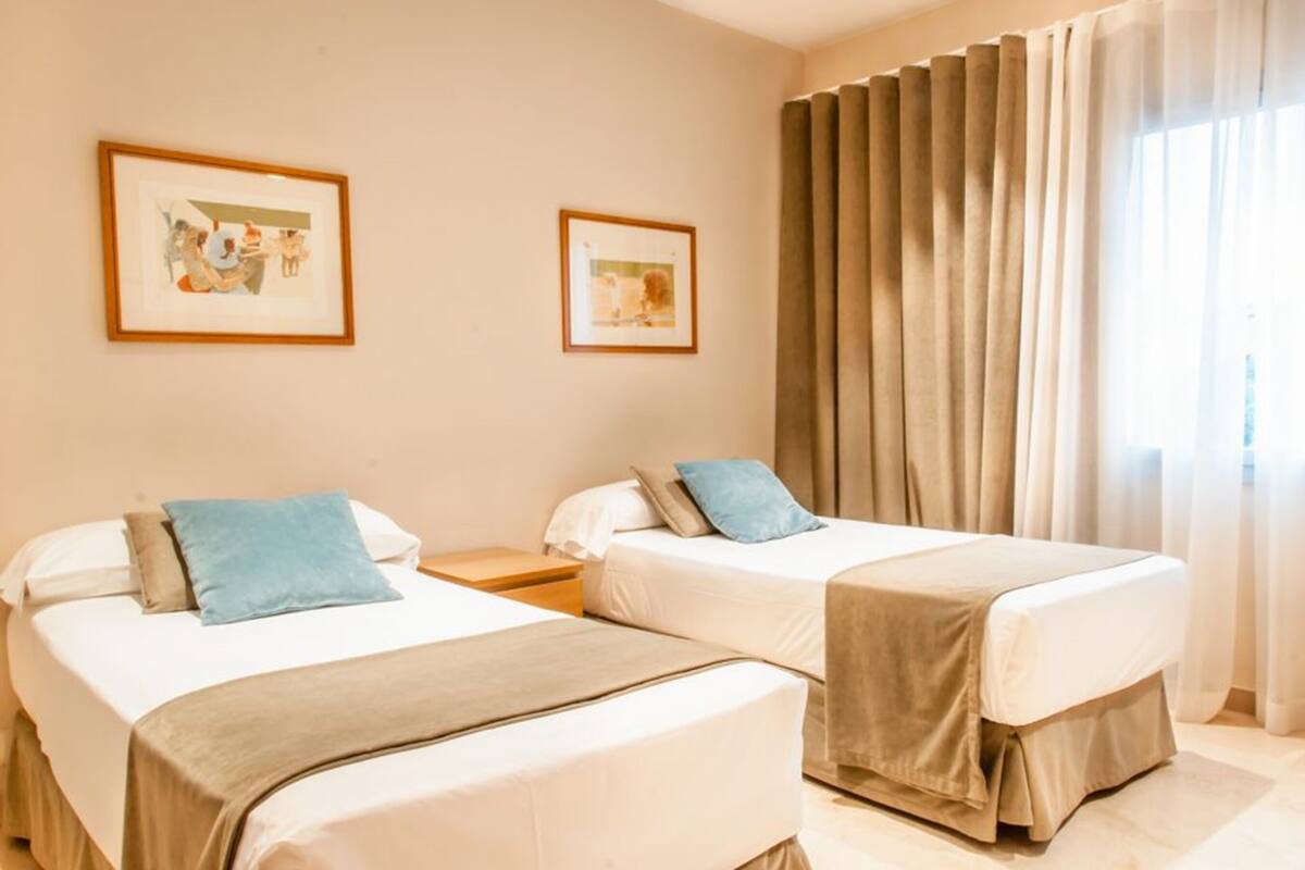 Quarto de hotel com duas camas de solteiro, uma mesa de cabeceira, um quadro na parede e uma janela com cortinas.
