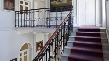 Uma escadaria imponente com grades ornamentadas e um candelabro, conduzindo a um andar superior com uma porta branca e um mural.