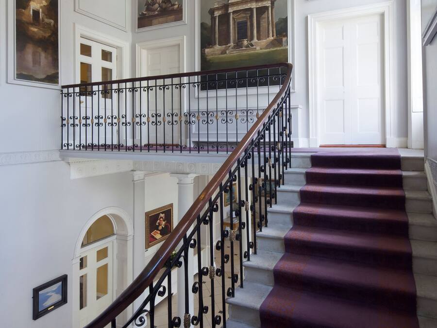 Uma escadaria imponente com grades ornamentadas e um candelabro, conduzindo a um andar superior com uma porta branca e um mural.