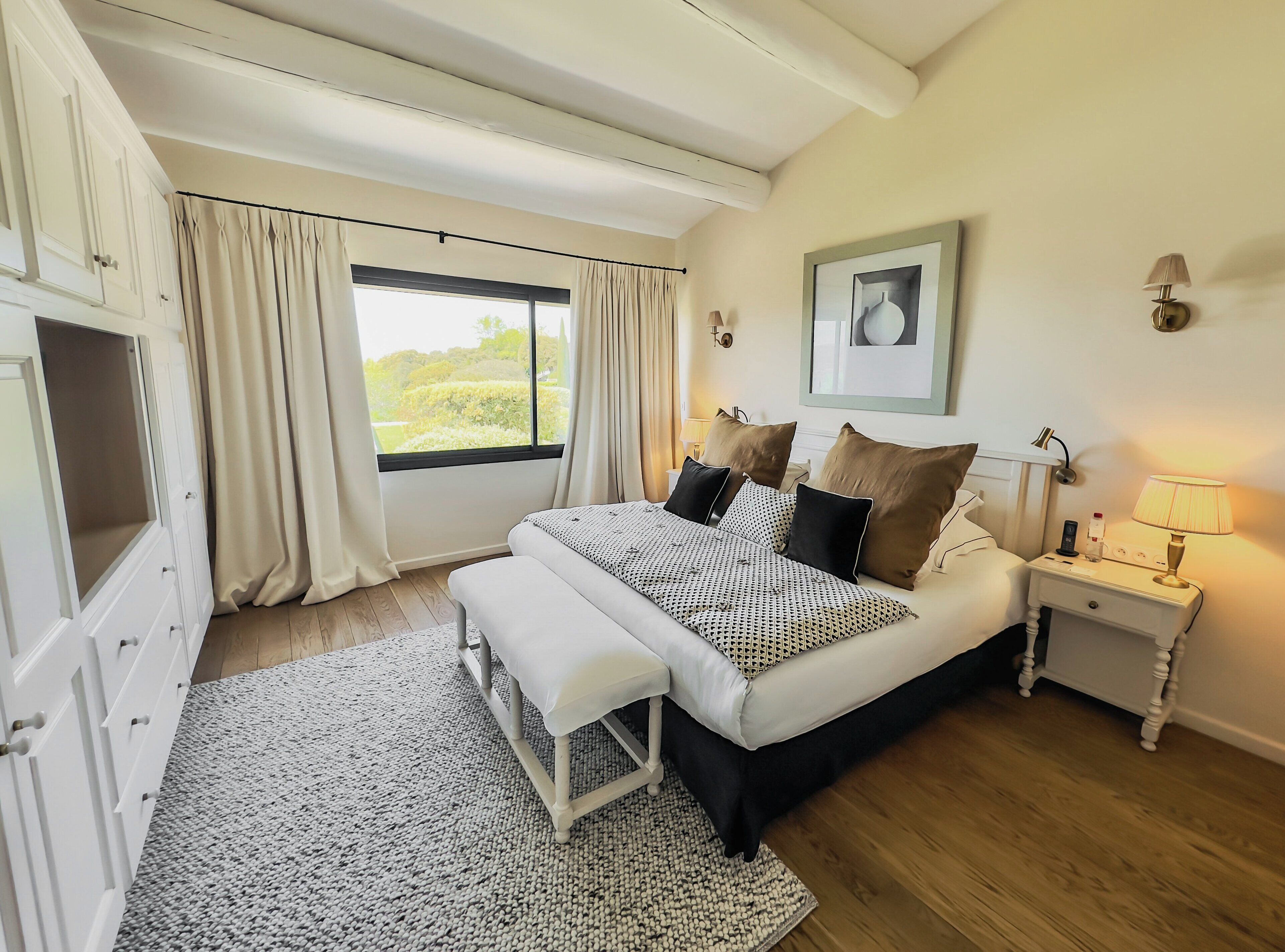 Quarto com cama grande, mesas de cabeceira, um banquinho, janela com cortinas e um quadro na parede.