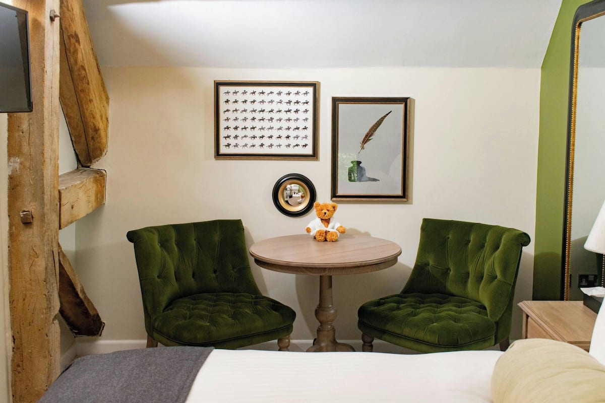 Quarto com cama, duas poltronas estofadas em verde, uma mesa de madeira redonda e duas molduras na parede.
