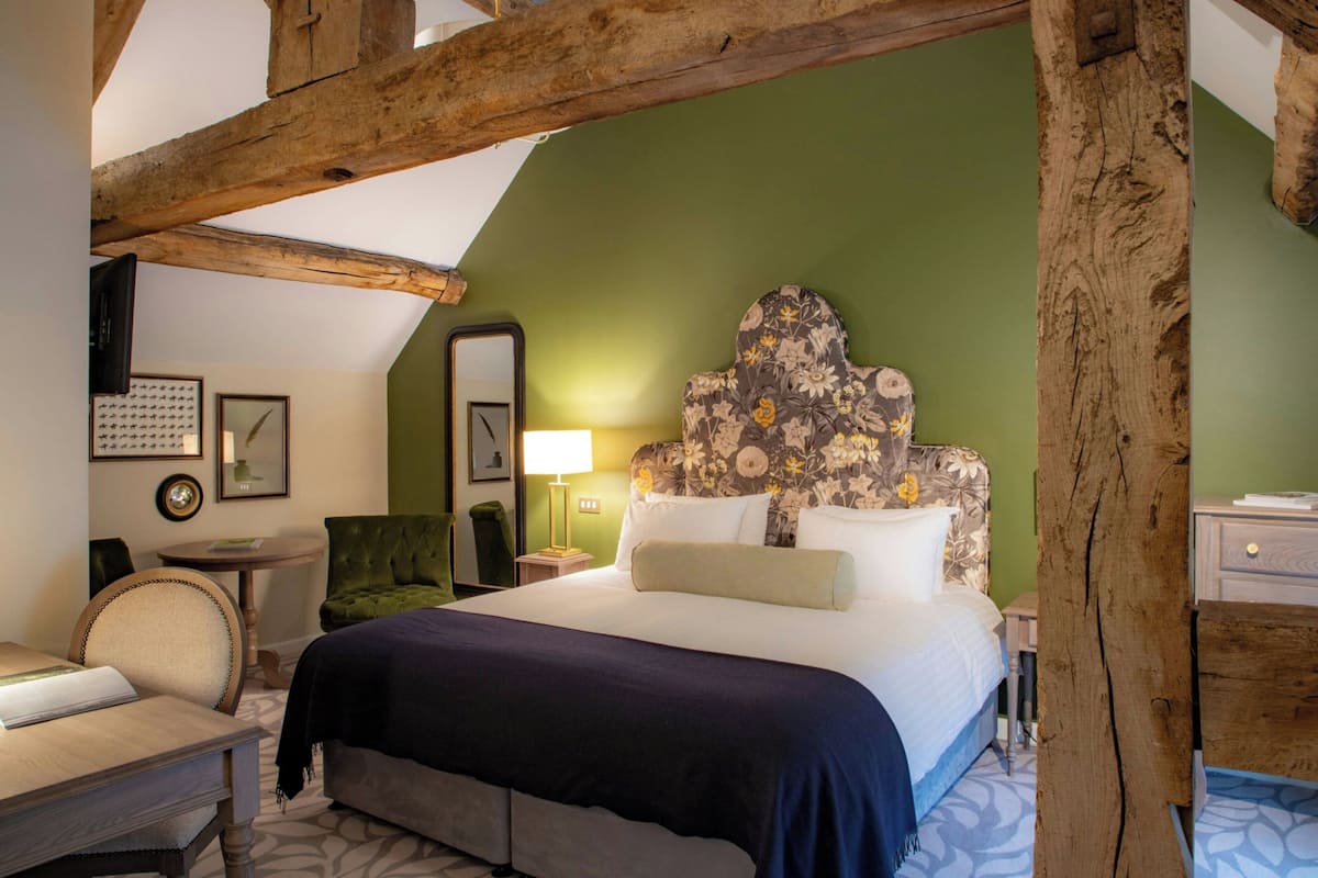 Quarto com uma cama grande, parede verde, vigas de madeira, poltrona verde, mesa pequena e luminária.