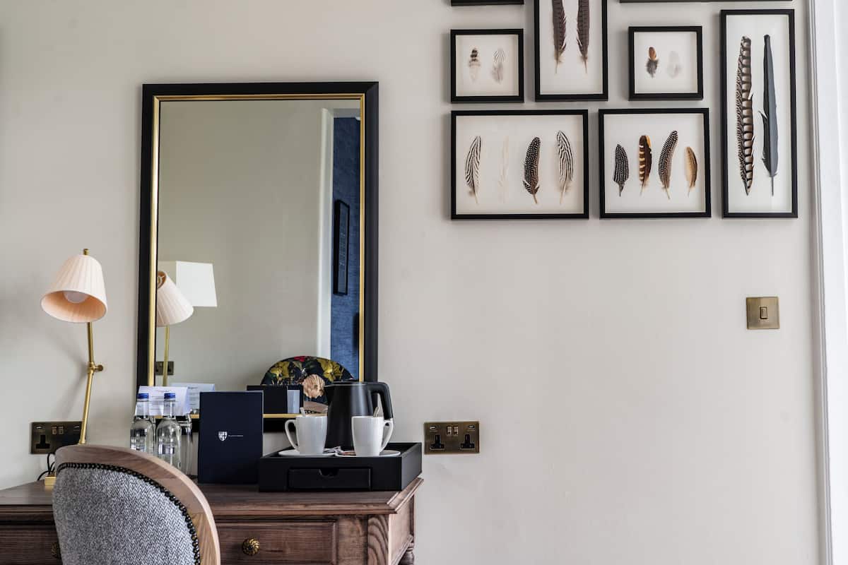 Quarto de hotel com um espelho grande, uma mesa com luminária, uma cafeteira e penas emolduradas na parede.