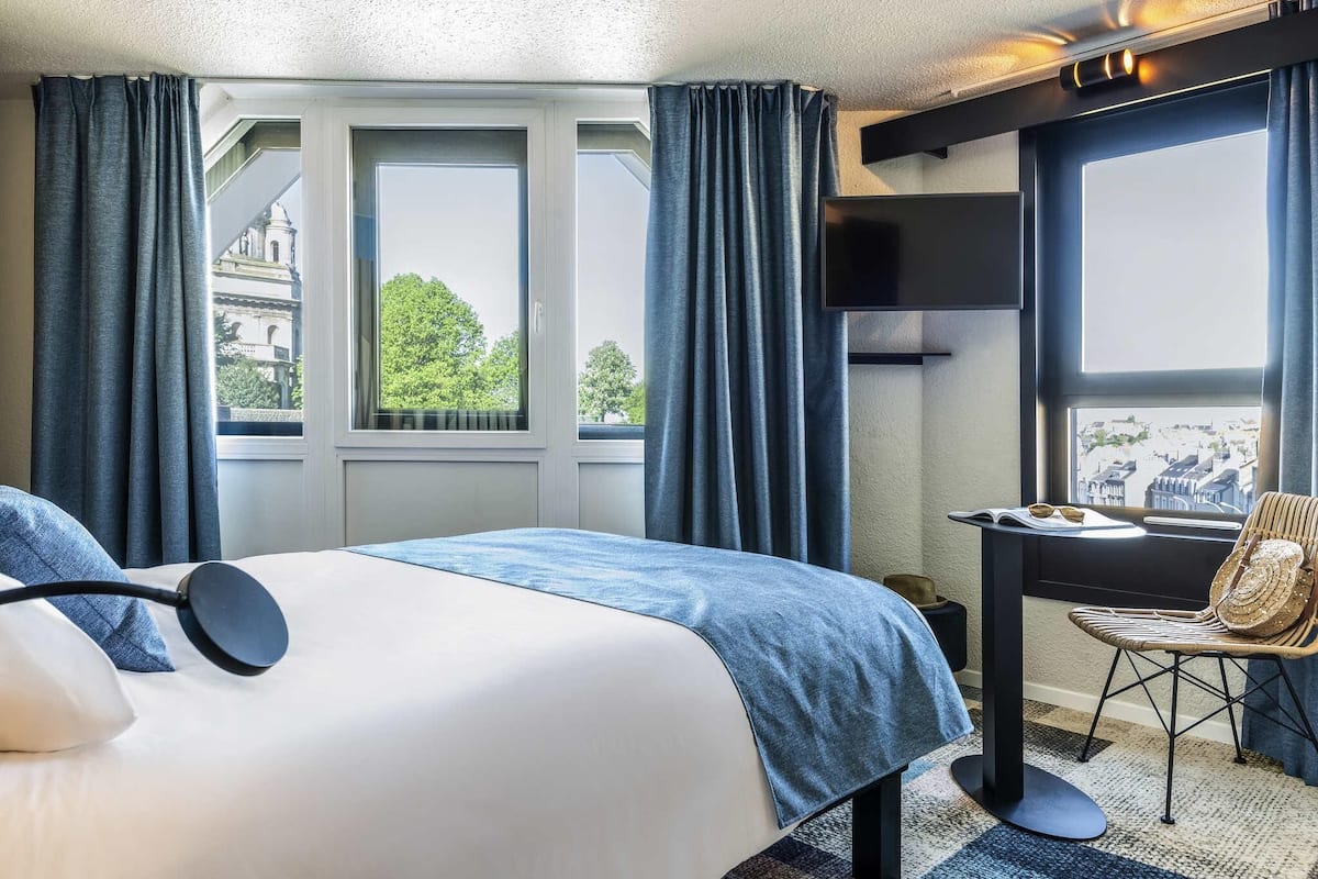 Quarto de hotel com cama, escrivaninha e cadeira. Há janelas com cortinas, vista de edifícios e paisagem urbana visível pelas janelas.