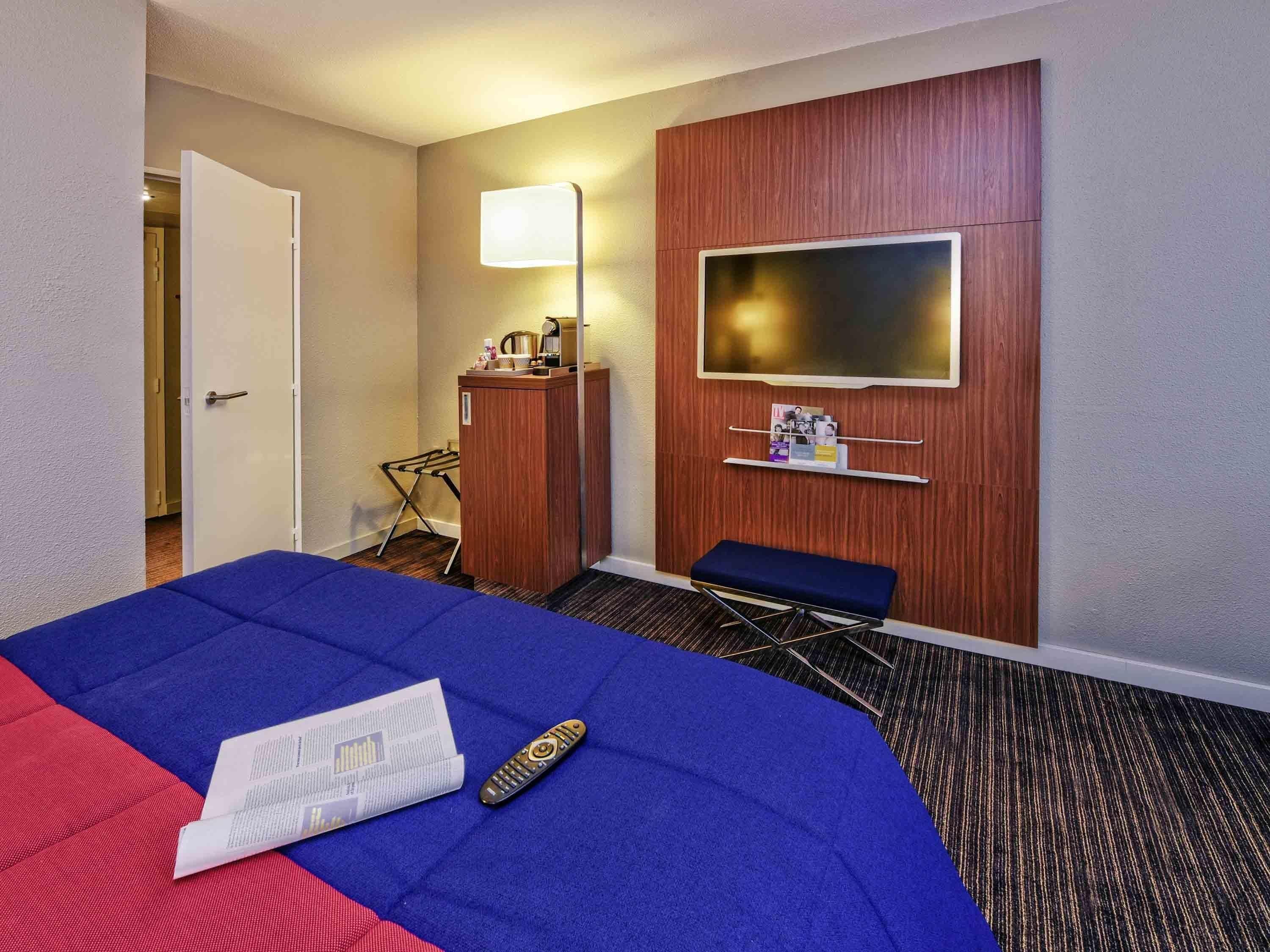 Quarto de hotel com uma cama grande, uma escrivaninha com luminária, televisão na parede e um pequeno banco.