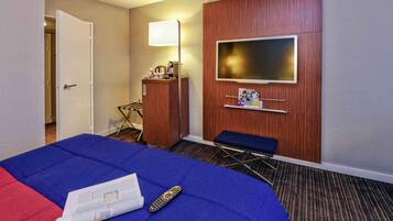 Quarto de hotel com uma cama grande, uma escrivaninha com luminária, televisão na parede e um pequeno banco.