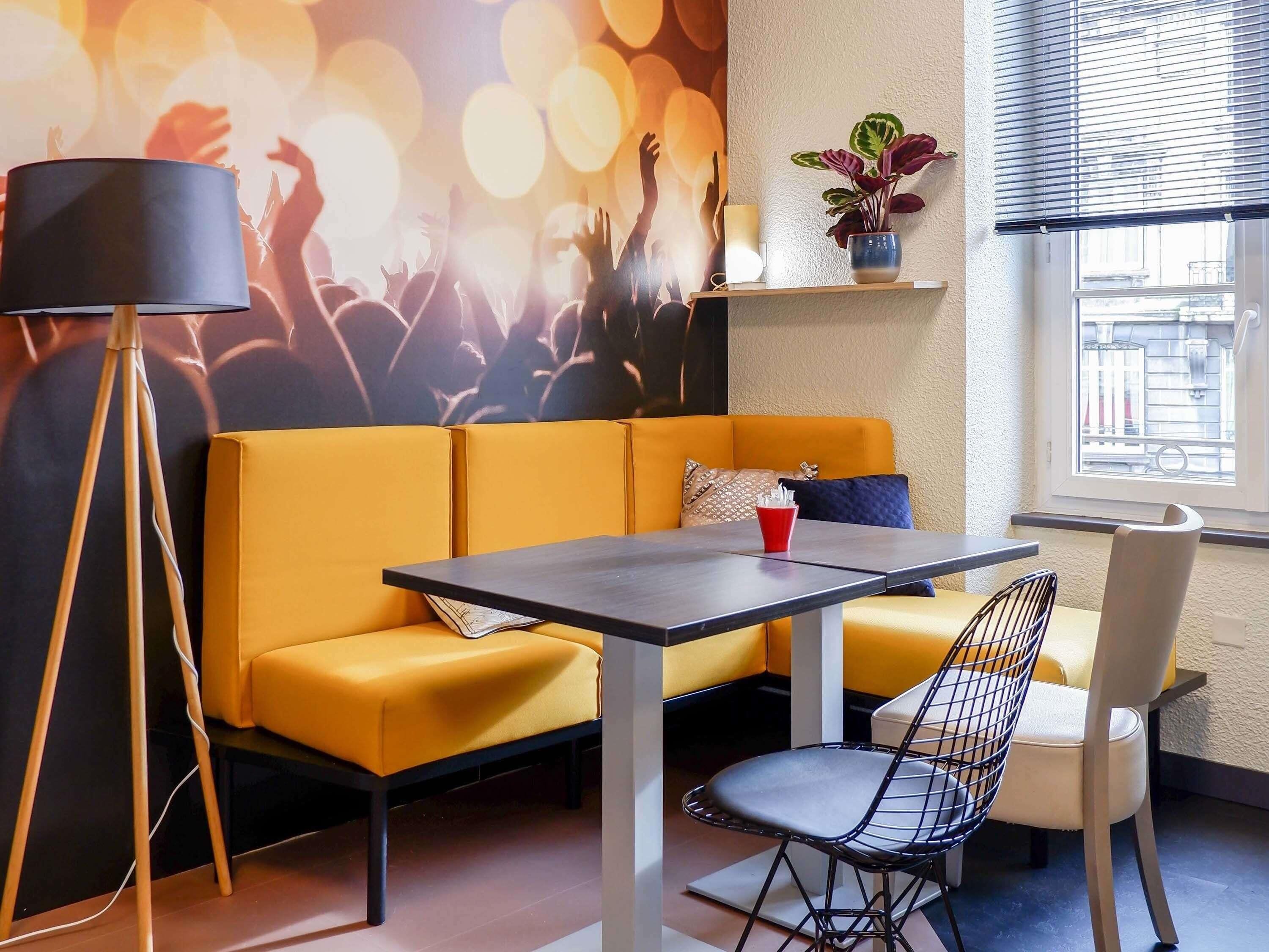 Um quarto com um sofá amarelo, uma mesa preta, uma cadeira de arame, um abajur e uma janela com persianas.