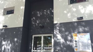 Entrada de um hotel com porta de vidro, placa indicando os serviços e vista para a rua e árvores.