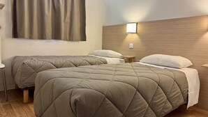 Quarto de hotel com duas camas, cabeceira de madeira, mesa de cabeceira, luminária e janela com cortinas.