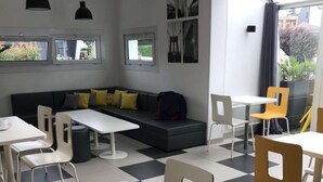 Quarto moderno e bem iluminado, com piso xadrez, cadeiras brancas e amarelas, um sofá cinza com almofadas amarelas e uma grande janela com vista para o exterior.