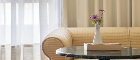Quarto de hotel com cama, sofá, mesa redonda com um vaso de flores e cortinas leves.