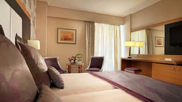Quarto de hotel com uma cama grande, duas cadeiras, uma mesa pequena com um vaso de flores, uma escrivaninha com abajur e uma televisão.