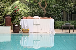 Uma pessoa deitada sobre uma toalha branca ao lado de uma piscina, com uma cesta de vime cheia de frutas e um vaso de flores por perto.