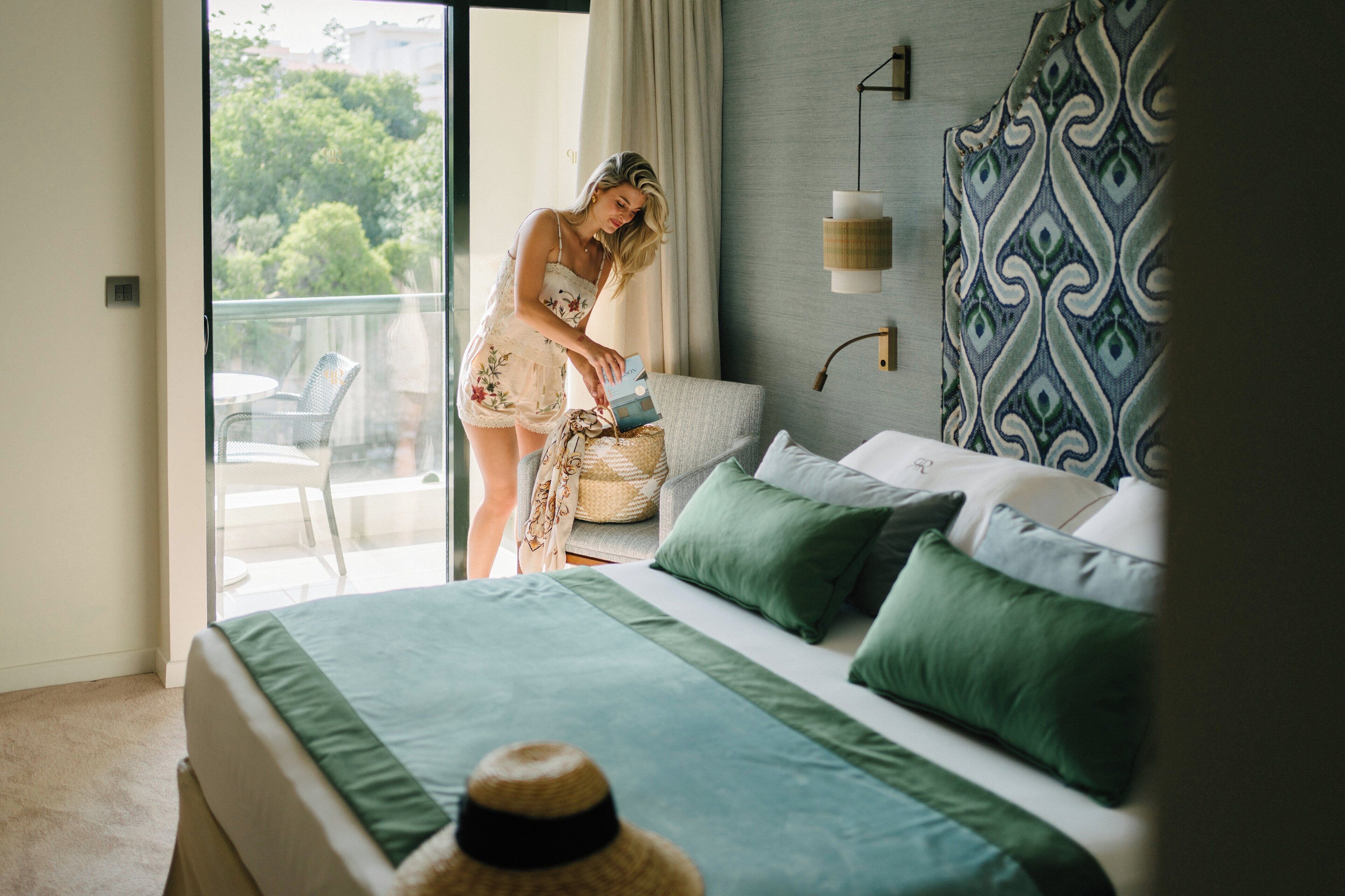 Quarto com cama, almofadas verdes, um chapéu na cama, varanda com cadeira e vista para árvores.