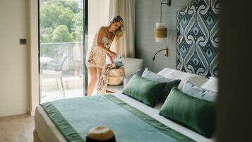 Quarto com cama, almofadas verdes, um chapéu na cama, varanda com cadeira e vista para árvores.