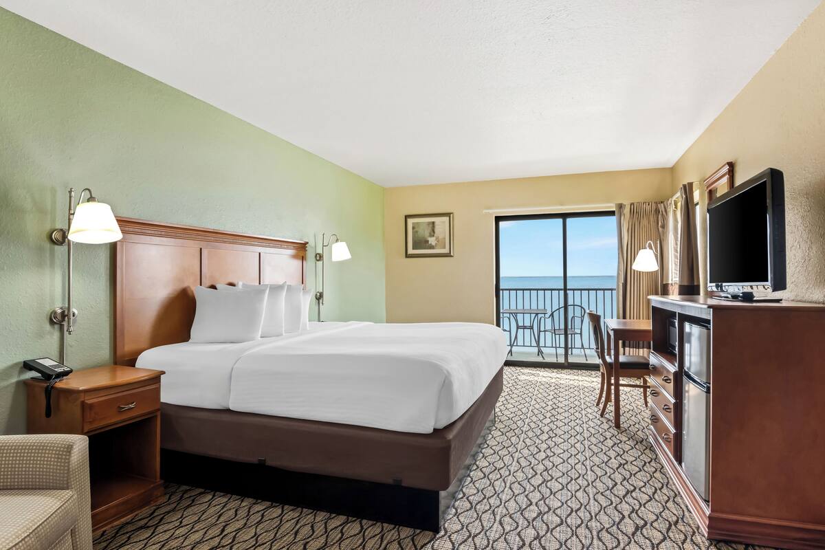 Quarto de hotel com cama, abajures, escrivaninha com telefone, cômoda, televisão e varanda com vista para o céu.