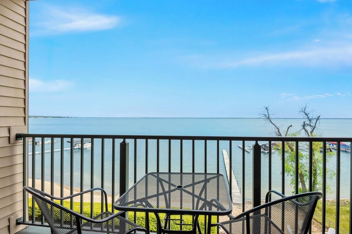 Uma varanda com mesa e cadeiras, com vista para um corpo de água calma, onde se vê um cais e barcos ao longe.