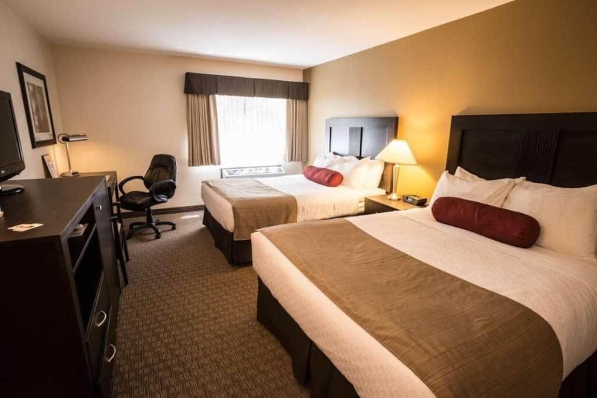 Quarto de hotel com duas camas, uma escrivaninha, uma cadeira, televisão e janela com cortinas.
