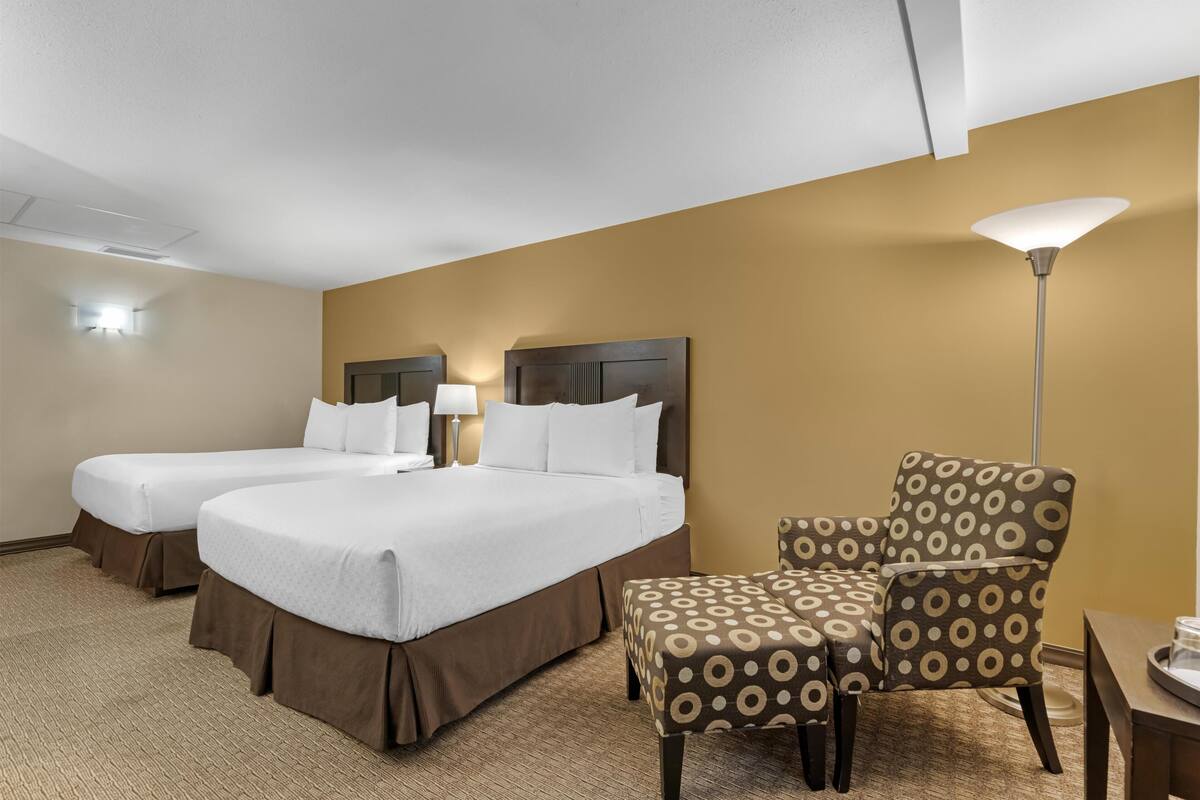 Quarto de hotel com duas camas, uma cadeira, um abajur e uma pequena mesa.