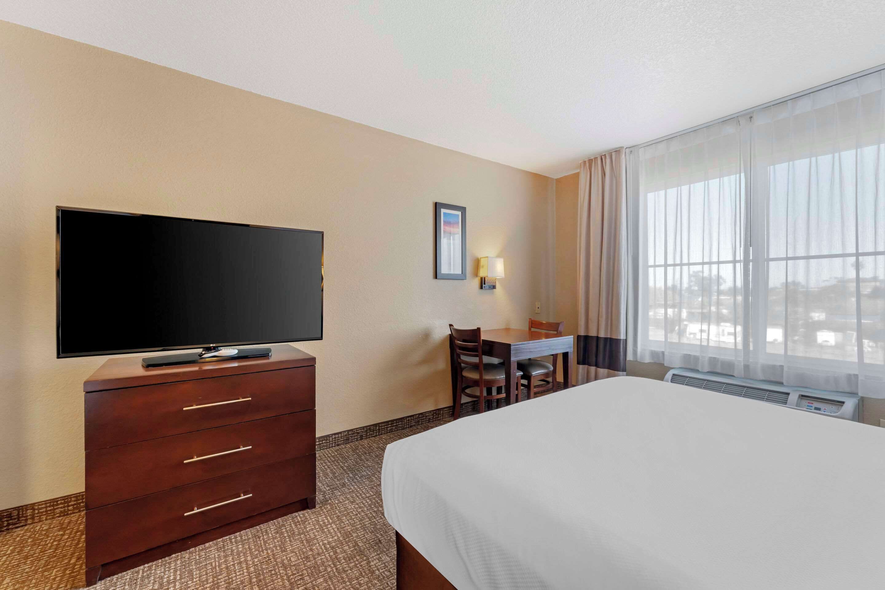 Quarto de hotel com cama, televisão em uma cômoda, escrivaninha com cadeira e uma grande janela com cortinas.